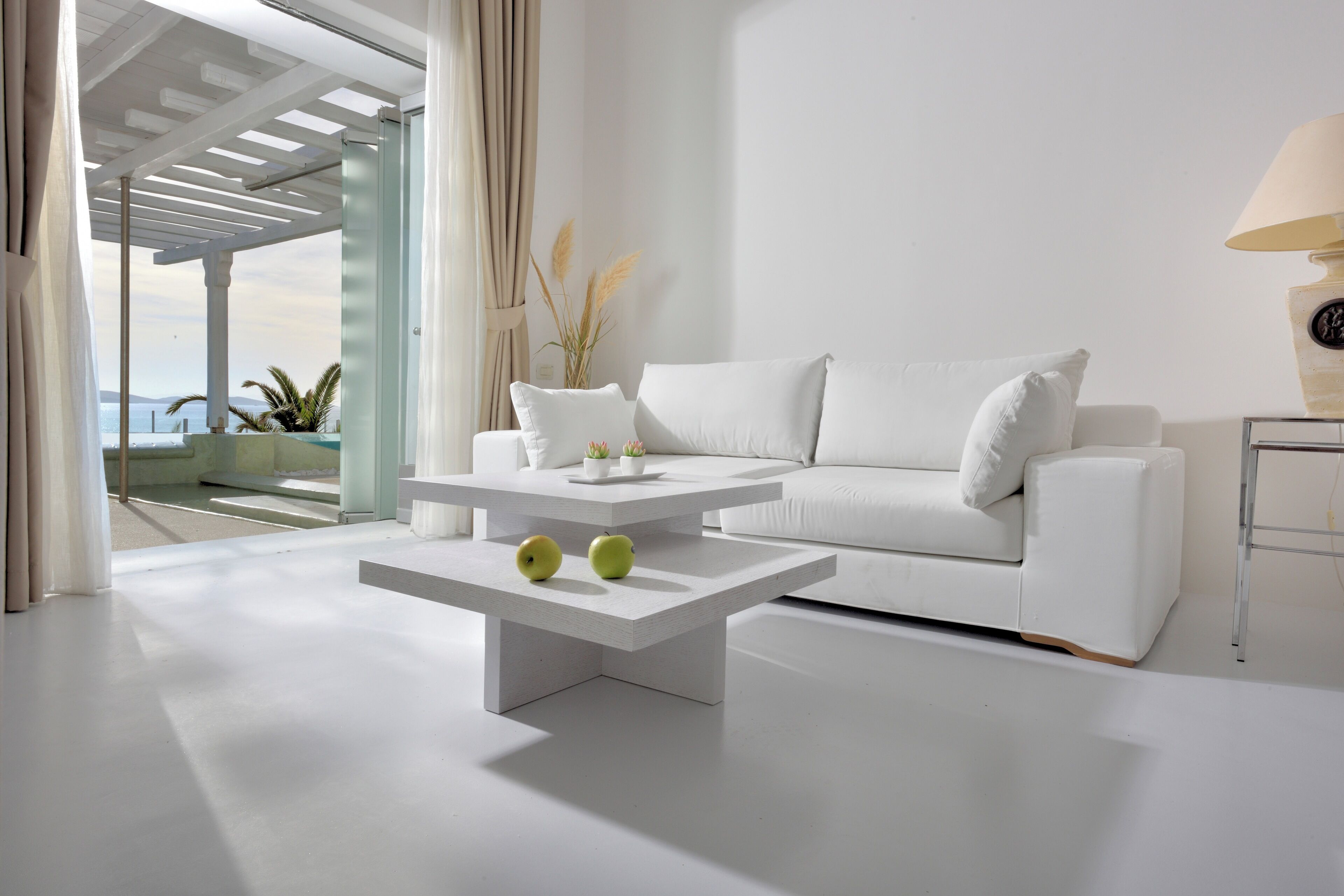 Uma sala de estar moderna com um sofá branco, uma mesa de centro branca e vista para o mar através de grandes janelas.