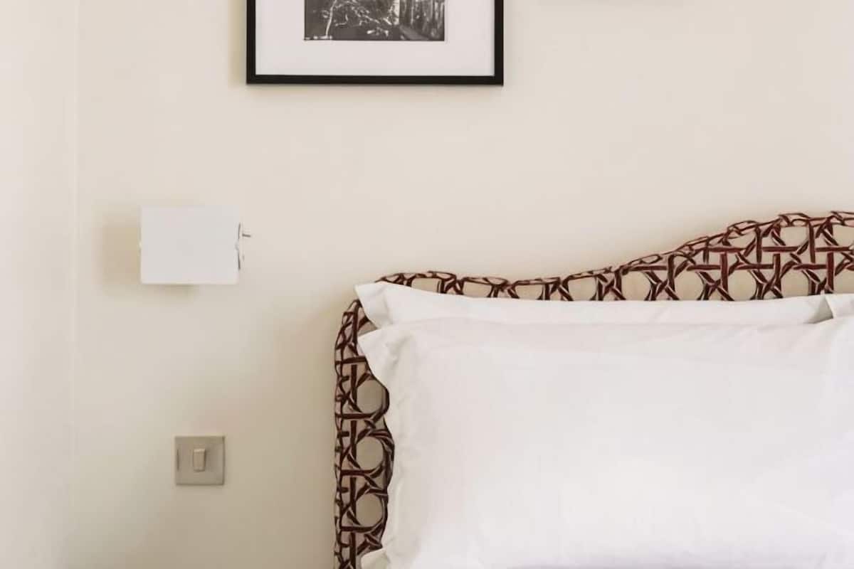 Cama com cabeceira estampada, travesseiro branco, quadro na parede e interruptor de luz.