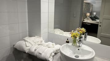 Um banheiro moderno com pia branca, porta-toalhas e espelho refletindo uma cama e mesa de cabeceira.