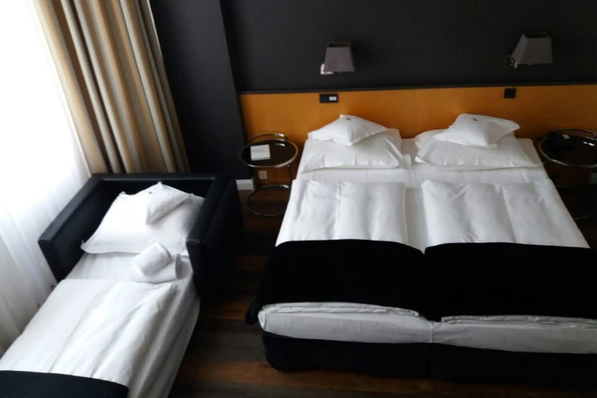 Quarto de hotel com duas camas, uma cabeceira de madeira, uma mesinha de cabeceira e uma janela com cortinas.