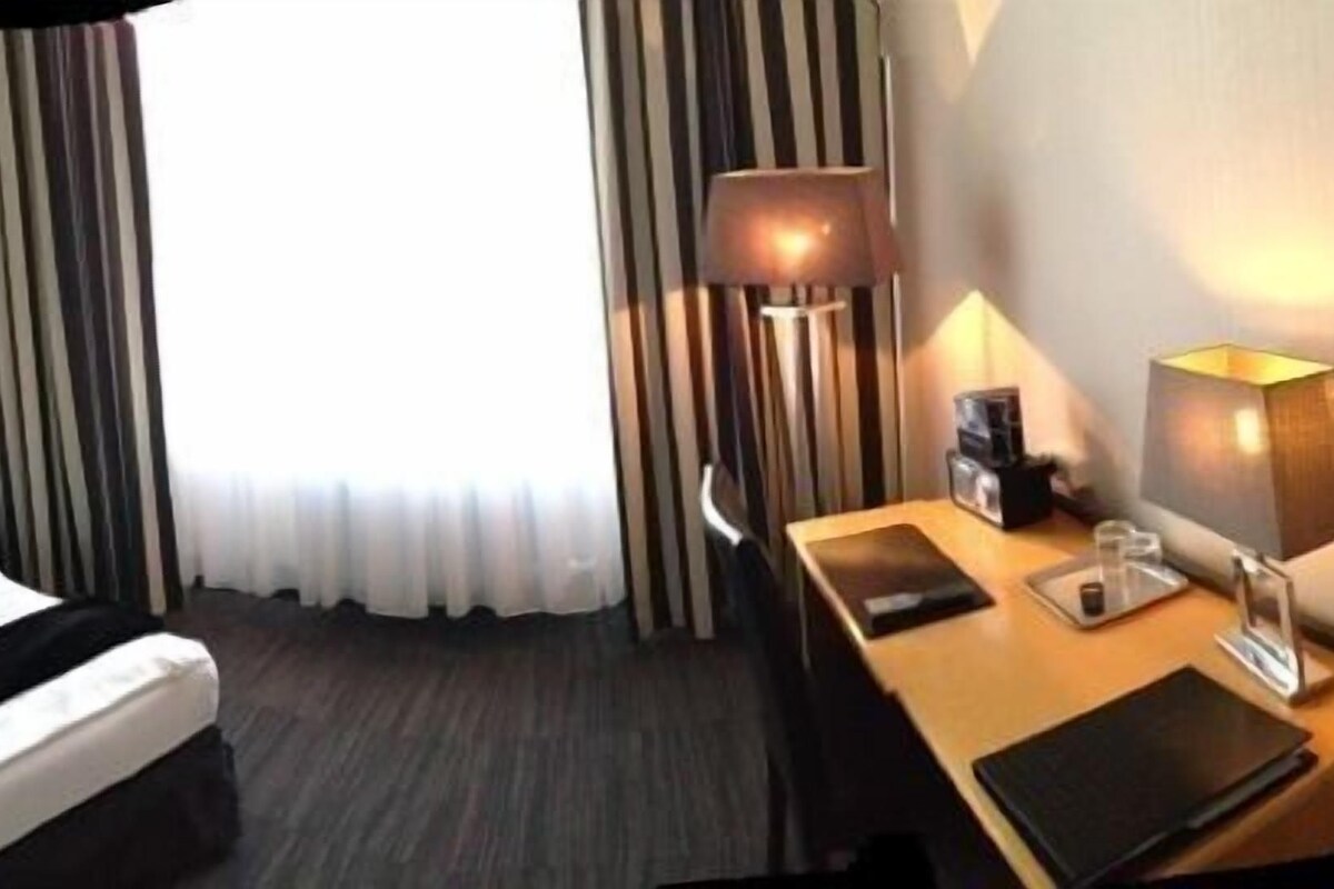 Quarto de hotel com cama, escrivaninha, luminária, televisão e janela com cortinas.