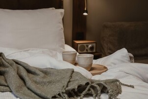 Cama com roupa de cama branca, um cobertor cinza e duas chávenas de cerâmica sobre uma bandeja de madeira.