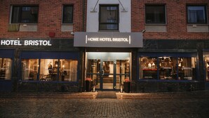 Um hotel com fachada de tijolos, uma placa com o texto 'HOME HOTEL BRISTOL' e grandes janelas que exibem os móveis do interior.