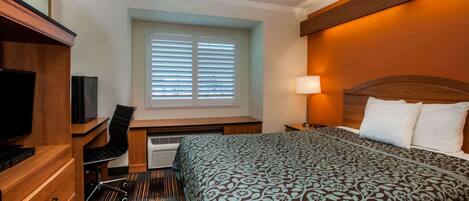 Quarto de hotel com cama, escrivaninha, cadeira, televisão e janela com persianas.