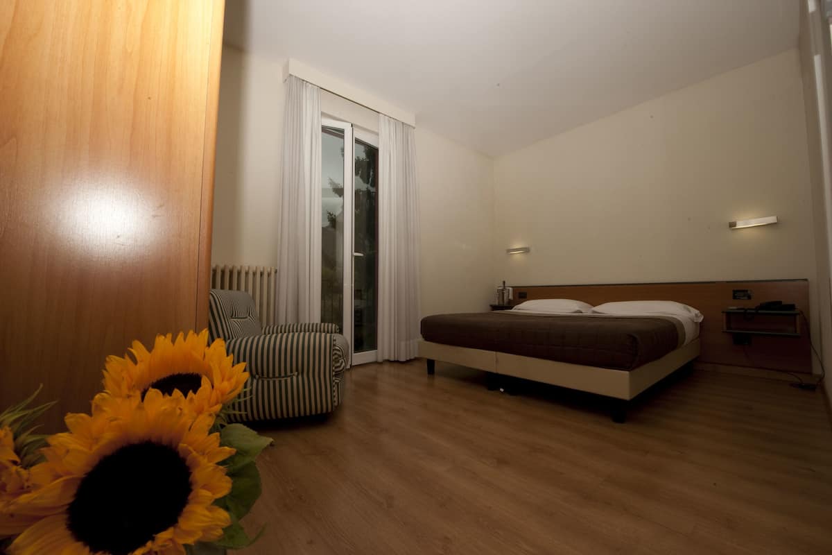 Quarto de hotel com piso de madeira, cama com cabeceira marrom, poltrona listrada, arranjo de girassóis e porta de vidro deslizante.