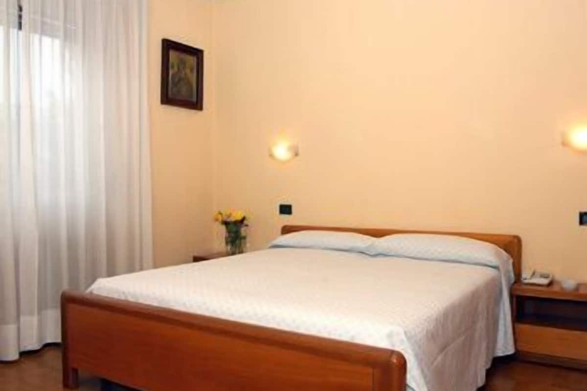 Quarto com cama de madeira, roupa de cama branca, mesa de cabeceira com abajur e iluminação embutida na parede.
