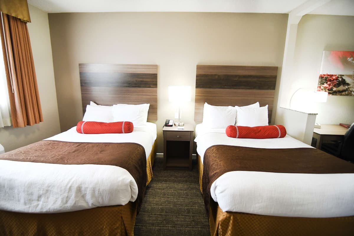 Quarto de hotel com duas camas, cada uma com cabeceira marrom, roupa de cama branca e almofadas vermelhas. Um criado-mudo entre as camas contém uma luminária e um telefone.