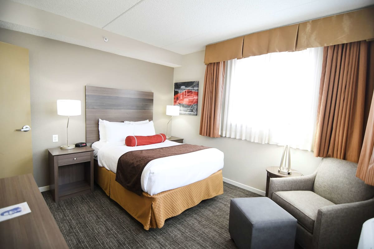 Quarto de hotel com cama, uma cadeira, mesa de cabeceira, luminária e janela com cortinas.