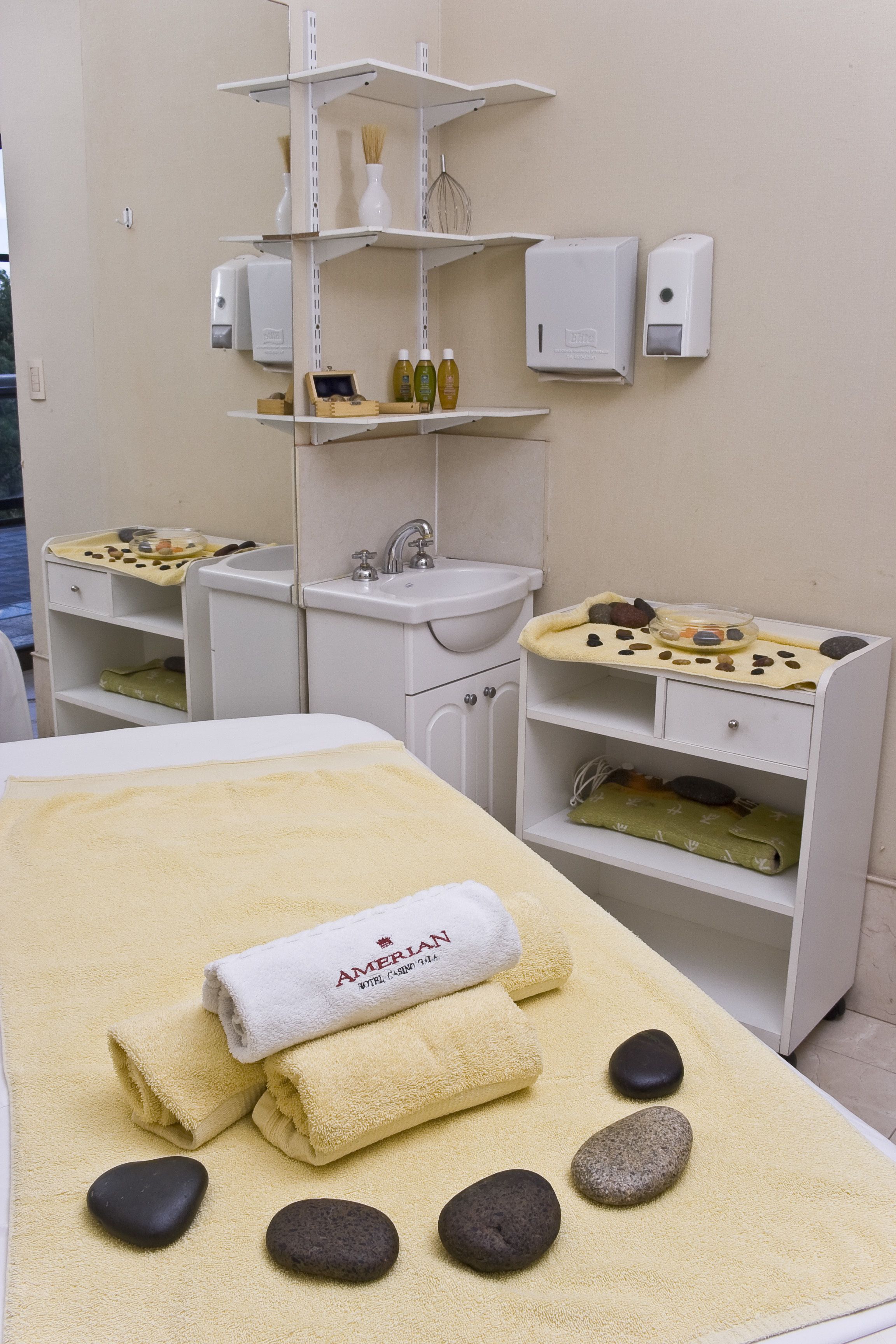 Uma sala de spa com mesa de massagem, toalhas e pedras. Há prateleiras com frascos e um dispensador de sabonete.
