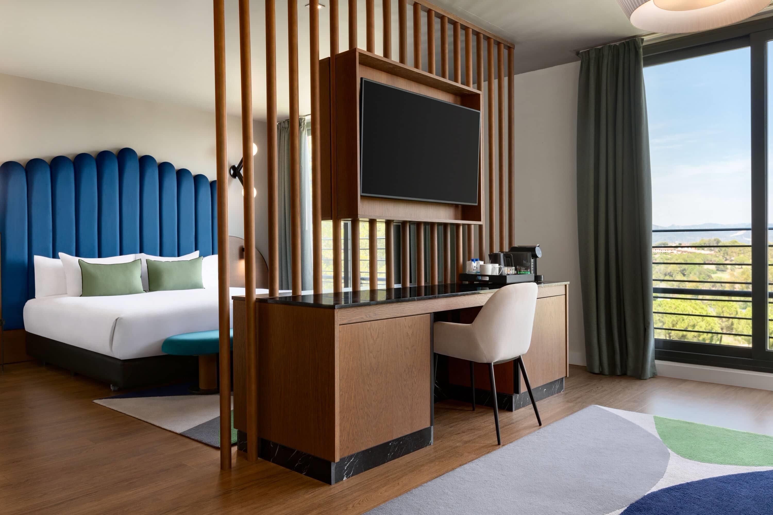 Quarto de hotel com cama, escrivaninha, cadeira, televisão e vista para o exterior.