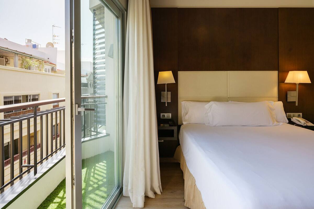 Quarto de hotel com varanda, cama com roupa branca, duas luminárias de cabeceira e mesa de cabeceira com telefone.