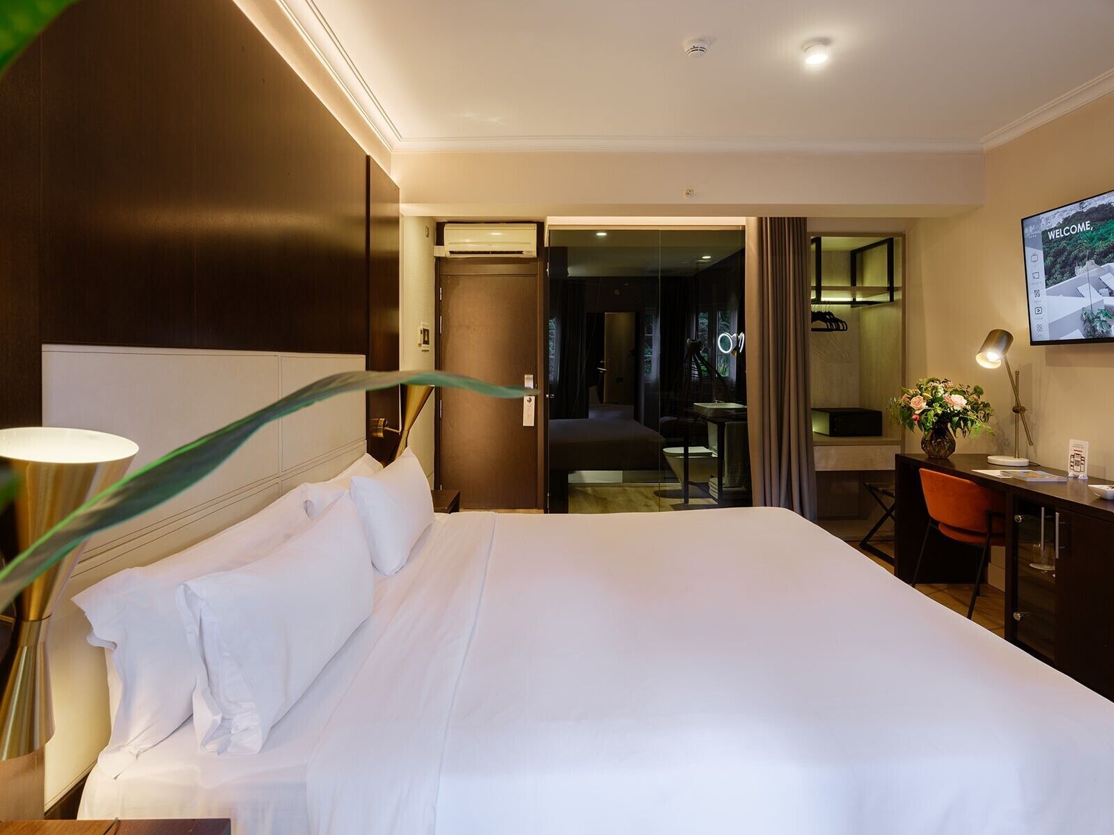 Quarto de hotel com uma cama grande, uma escrivaninha com cadeira, abajur, vaso com flores e televisão na parede.