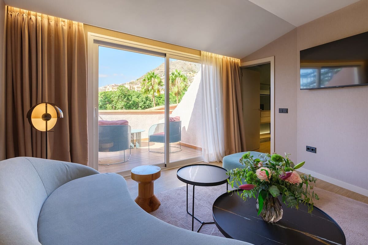 Quarto de hotel moderno com sofá curvo, uma pequena mesa redonda com um vaso de flores, luminária de chão e uma grande porta de vidro deslizante que dá para uma área externa com mobiliário e vista para vegetação e colinas.