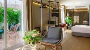 Quarto de hotel moderno com uma janela grande, uma cama, área de estar com uma poltrona, uma pequena mesa com um vaso de flores e um guarda-roupa suspenso.