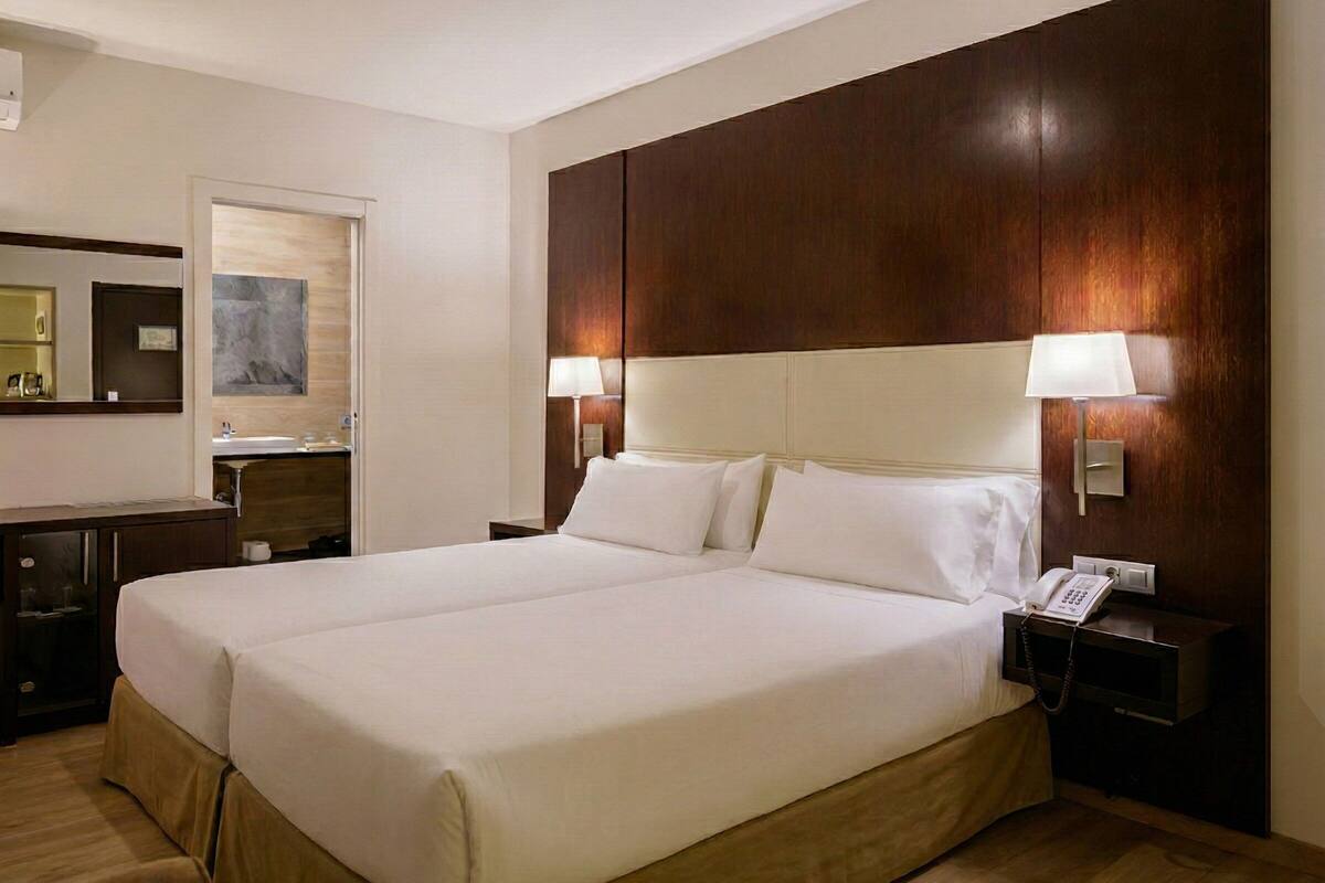 Quarto de hotel com uma cama grande, cabeceira de madeira, luminárias de cabeceira, telefone, espelho e mesa de cabeceira de madeira.