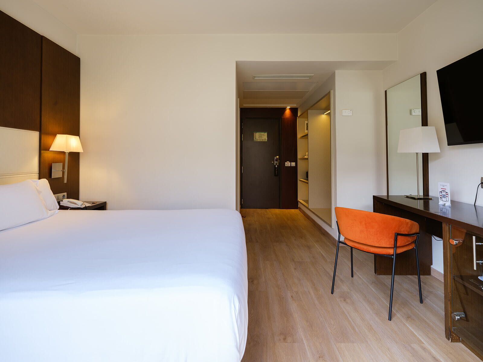Quarto de hotel com cama, escrivaninha com cadeira, televisão e porta para outro cômodo.