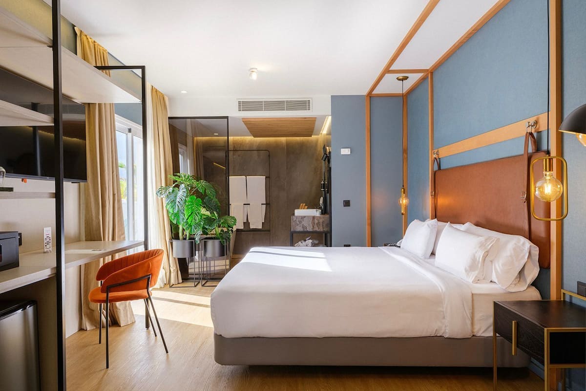 Quarto de hotel moderno com uma cama grande, uma escrivaninha, uma cadeira e um banheiro visível através de um painel de vidro.