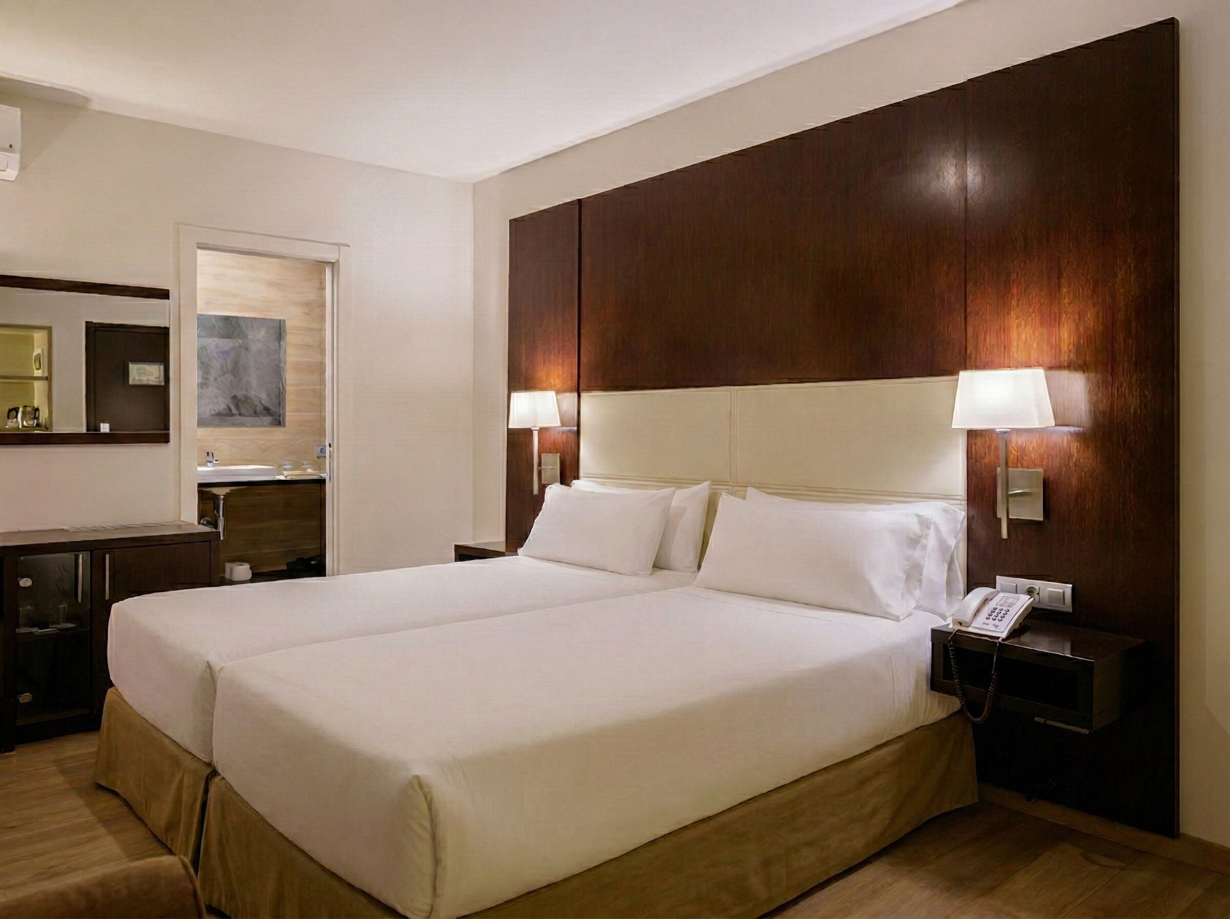 Quarto de hotel com uma cama grande, cabeceira de madeira, luminárias de cabeceira, telefone, espelho e mesa de cabeceira de madeira.