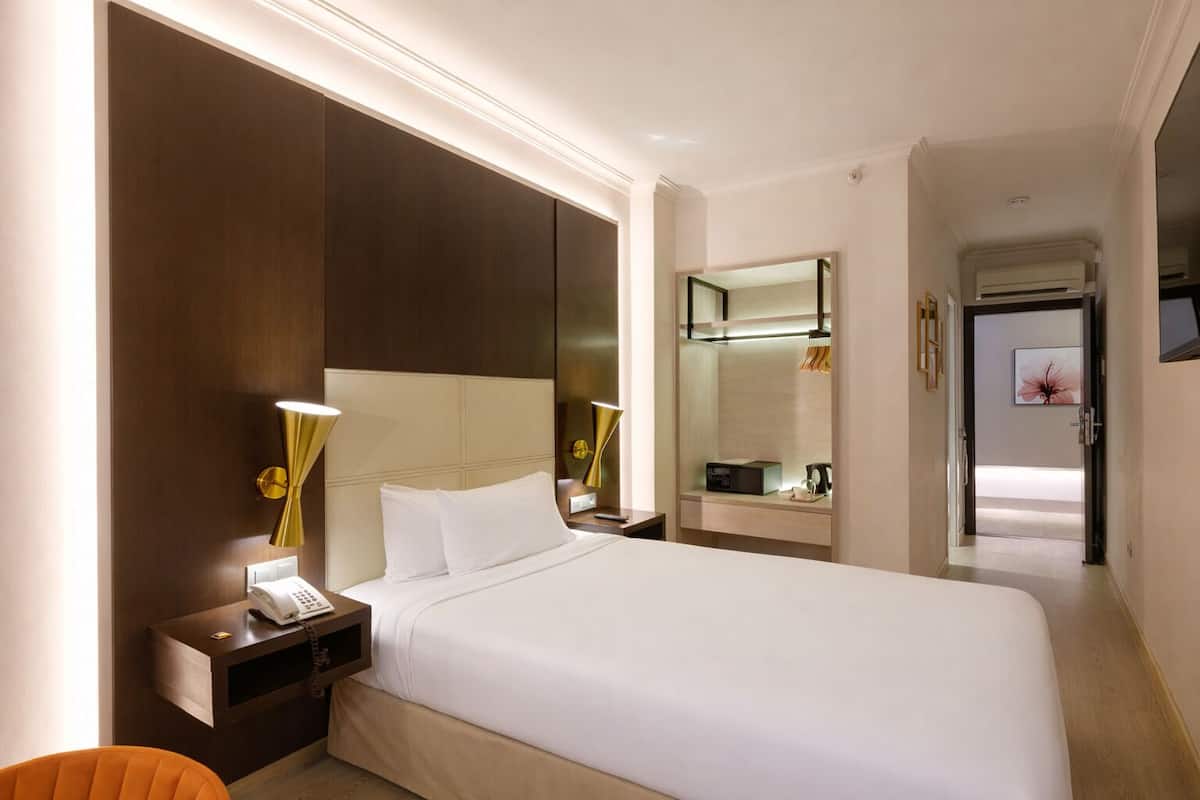 Quarto de hotel com uma cama grande, luminárias de cabeceira, mesa de apoio, espelho e uma porta que dá para outro cômodo.