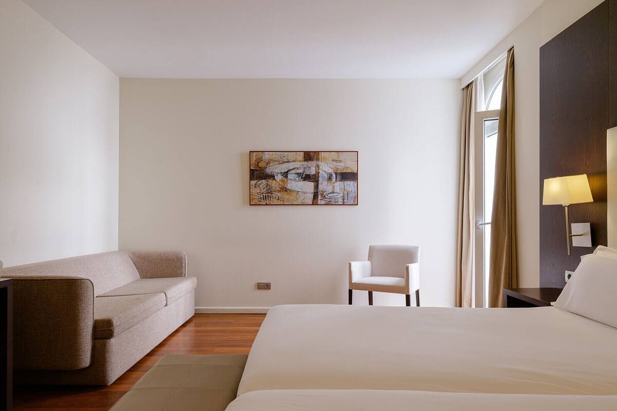 Quarto de hotel com cama, sofá, poltrona, quadro na parede e janela com cortinas.
