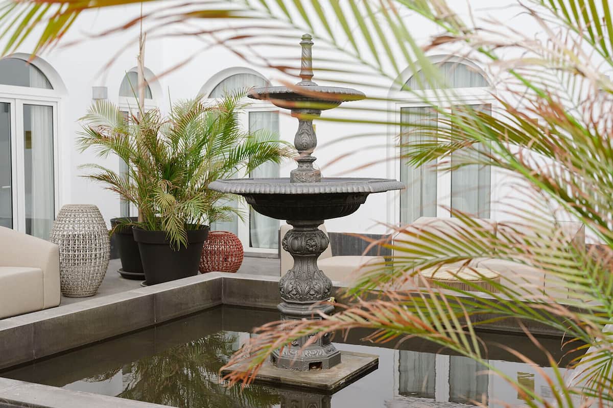 Uma fonte em um pátio com plantas em vasos e um edifício branco ao fundo.