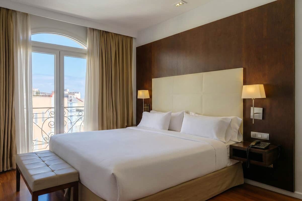 Quarto de hotel com uma cama grande, um banco, uma mesa de cabeceira com abajur e vista para a cidade através de uma janela ampla.