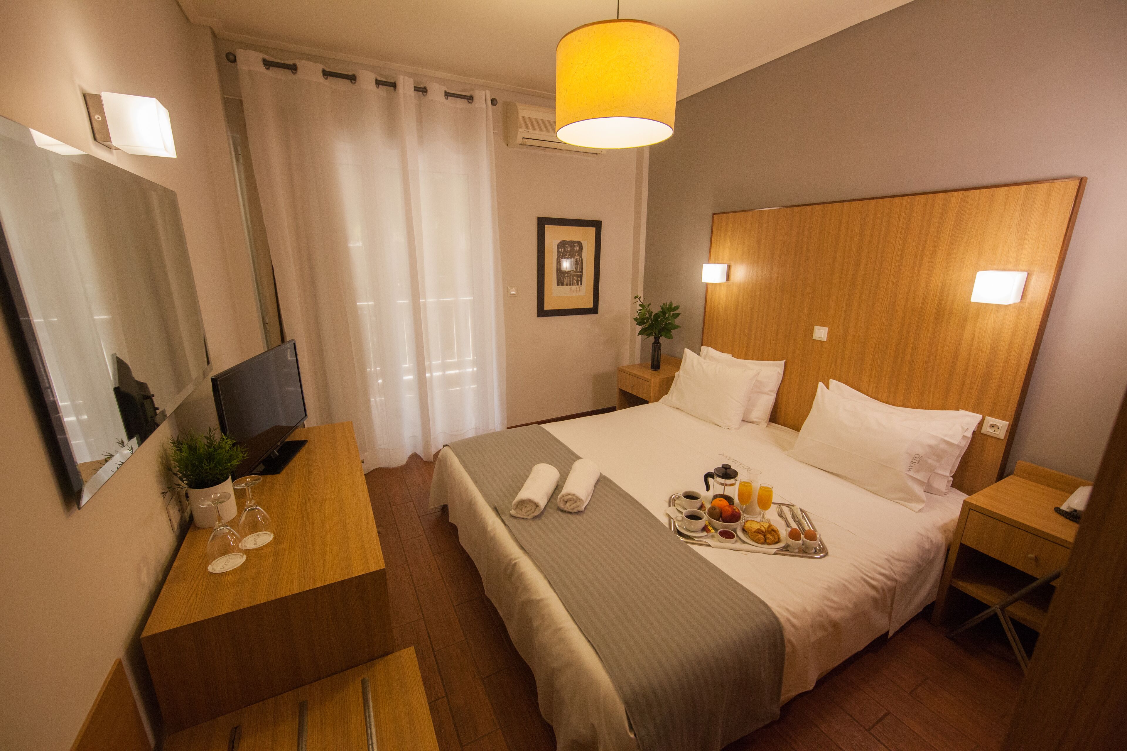Quarto de hotel com uma cama grande, uma escrivaninha de madeira com uma TV, uma mesinha de cabeceira com uma planta e uma bandeja de café da manhã na cama.