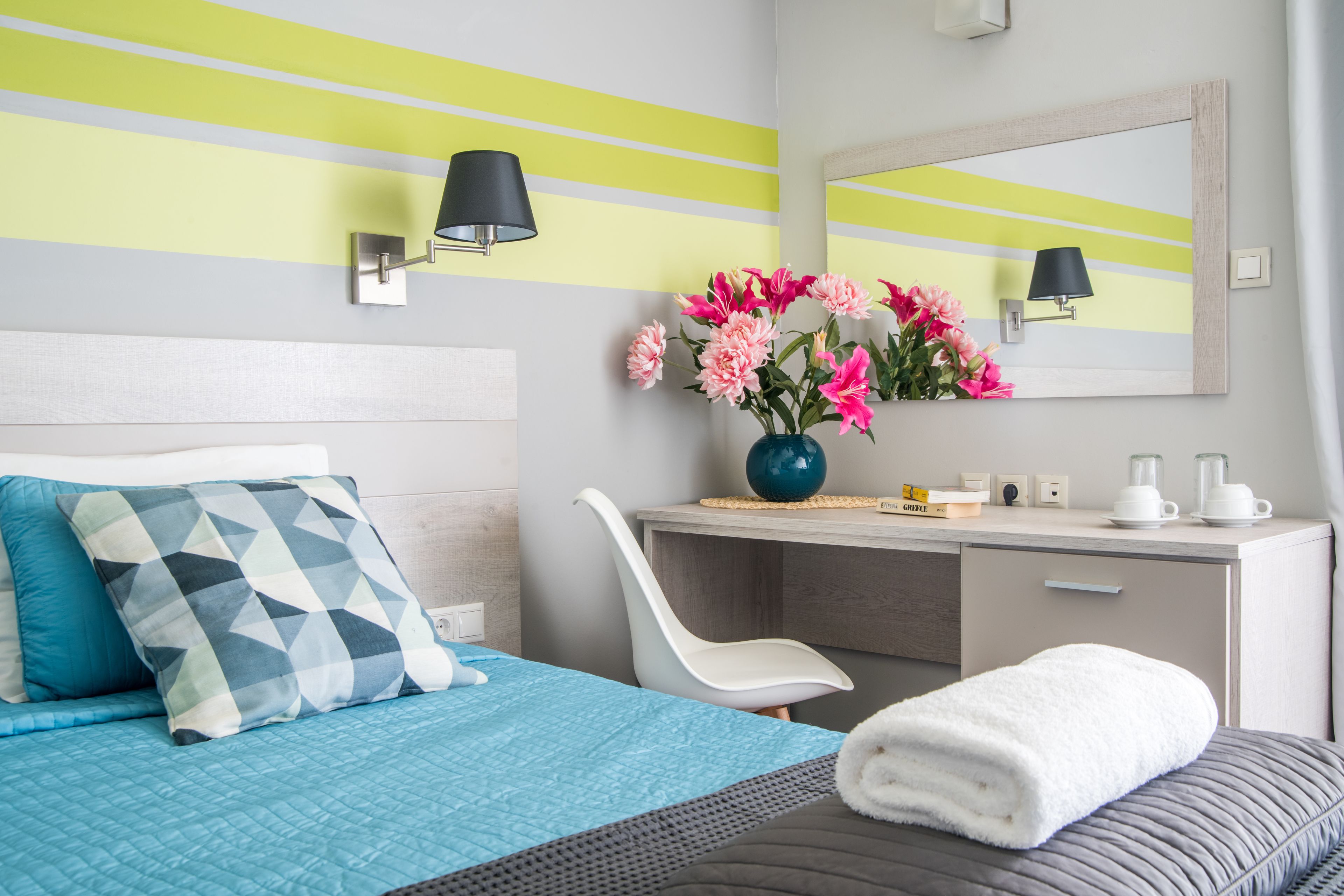 Quarto de hotel com cama, escrivaninha, cadeira, vaso com flores e espelho.