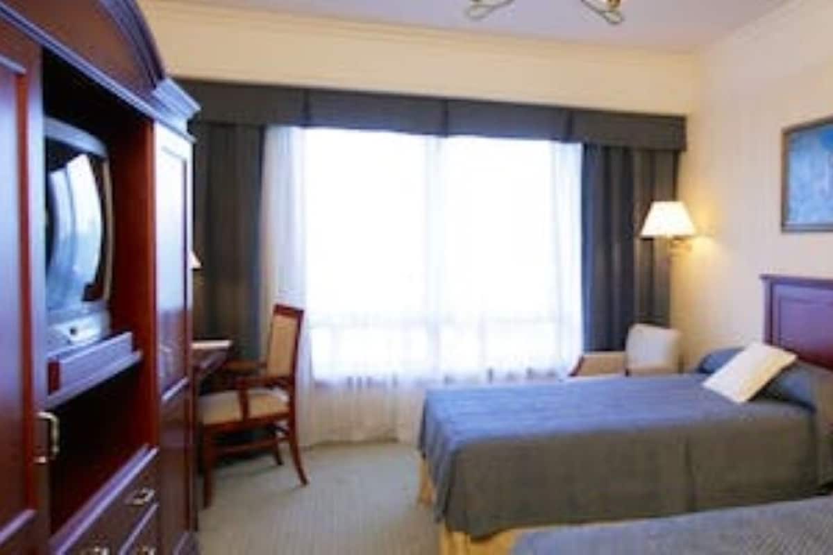 Quarto de hotel com duas camas, uma escrivaninha, uma cadeira, uma televisão e uma janela com cortinas.