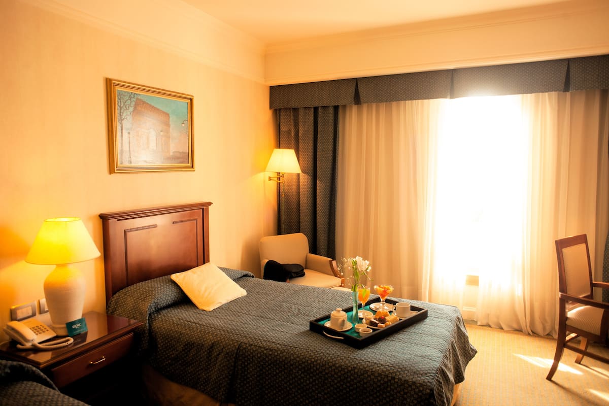 Quarto de hotel com cama, mesa de cabeceira com abajur, bandeja com itens de café da manhã, uma cadeira e um quadro na parede.