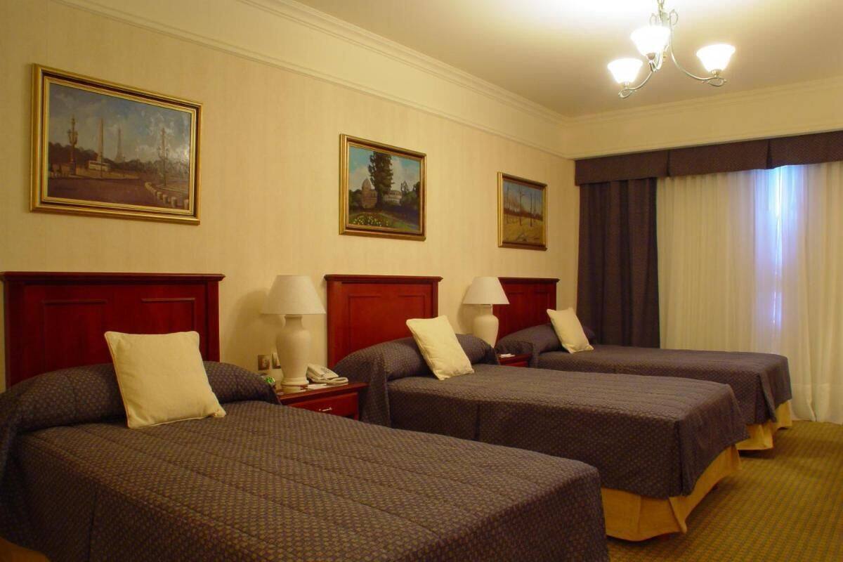 Quarto de hotel com três camas, cada uma com cabeceira vermelha, mesa de cabeceira com abajur e um quadro na parede.