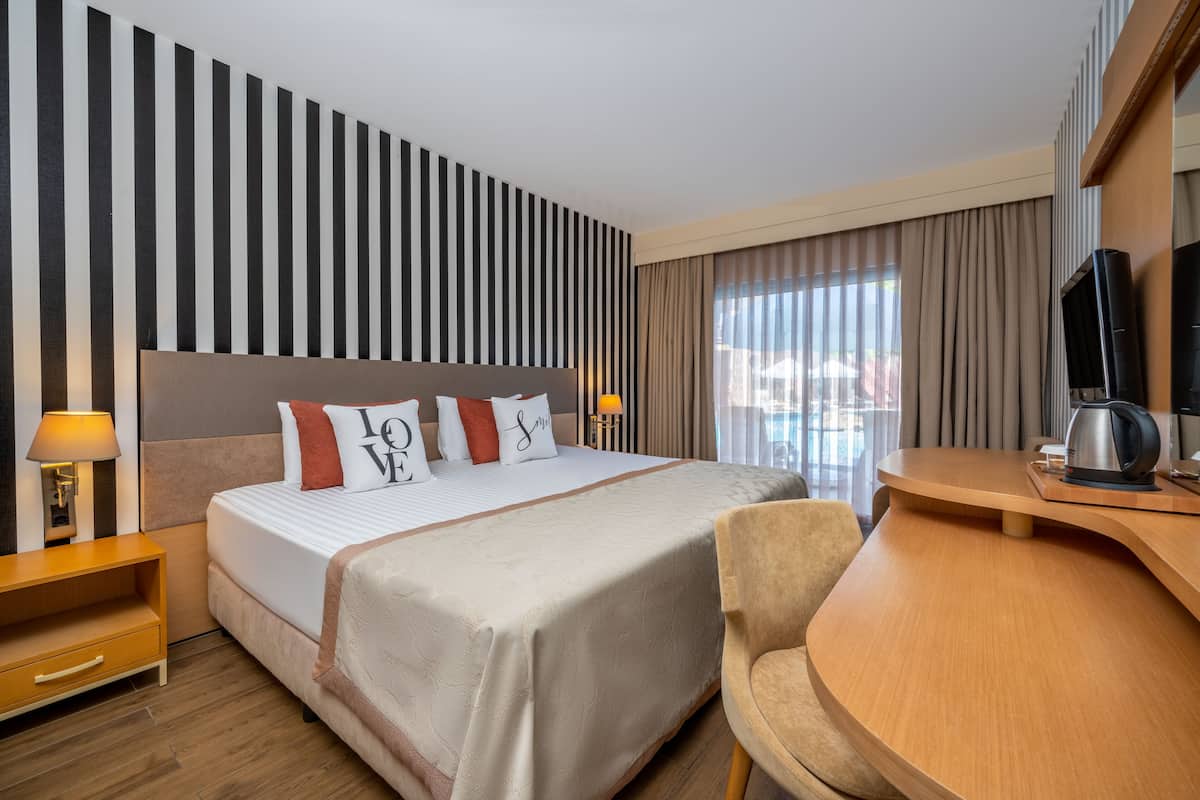 Quarto de hotel com cama, mesa de cabeceira, abajur, televisão e escrivaninha com cadeira.