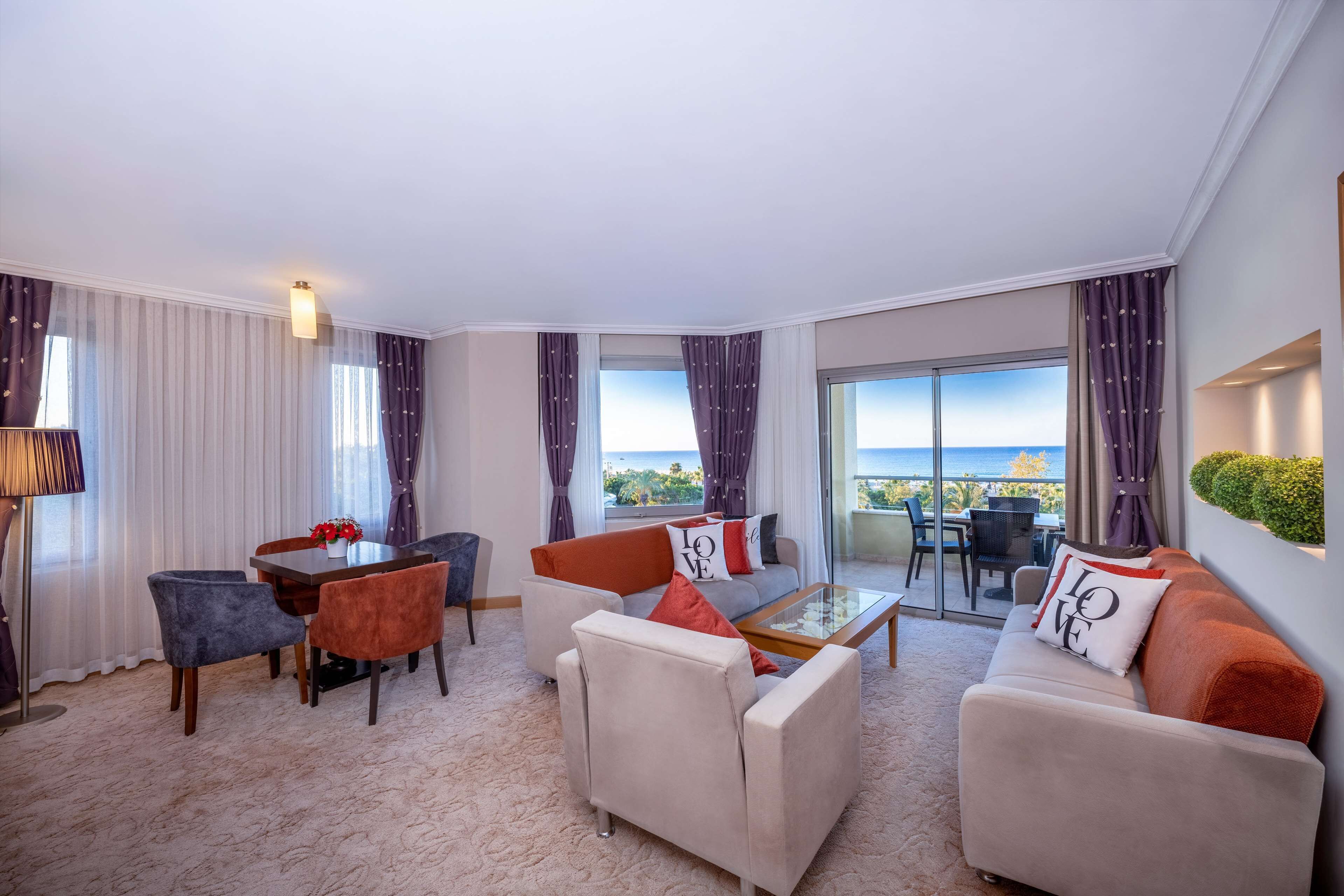 Uma sala de estar moderna com sofá, poltronas, mesa de centro e mesa de jantar. Há uma janela grande com cortinas, uma varanda com móveis de exterior e vista para o mar.