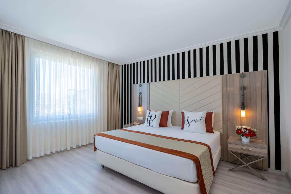 Quarto de hotel com uma cama grande, mesa de cabeceira com abajur, e vista para o exterior através de cortinas translúcidas.