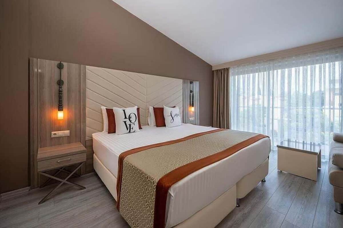Quarto de hotel moderno com uma cama grande, uma mesa de cabeceira e vista da cidade através de amplas janelas.