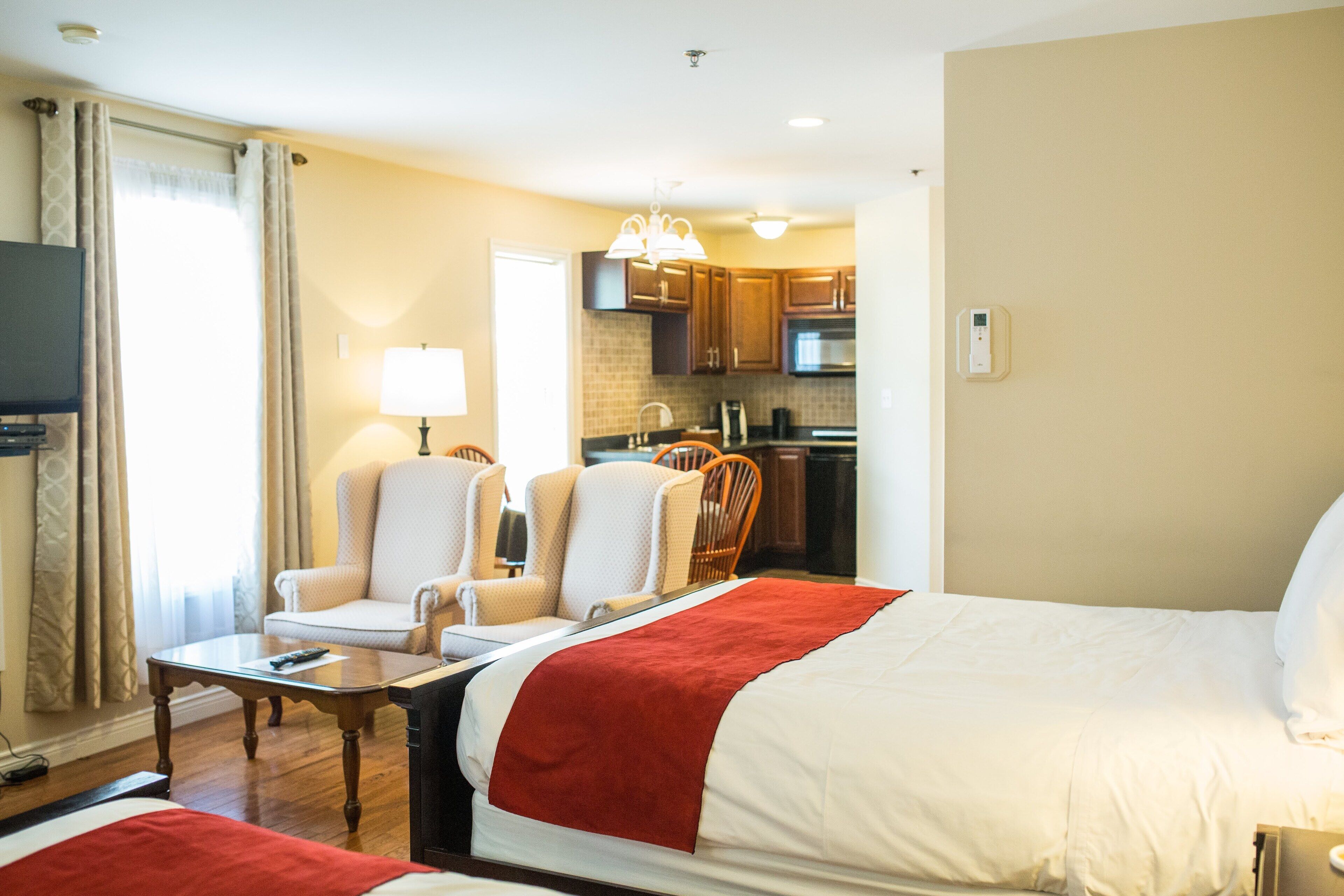 Quarto de hotel com cama, duas poltronas, uma mesinha, televisão e uma kitchenette ao fundo.