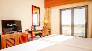 Quarto de hotel com televisão exibindo "VIP Executive Santa Iria Hotel", uma secretária com cadeira, uma lâmpada e uma janela grande com vista.