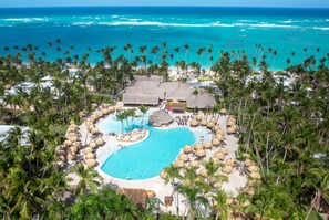 Vista aérea de um resort com piscina central rodeada por palmeiras e construções de colmo.