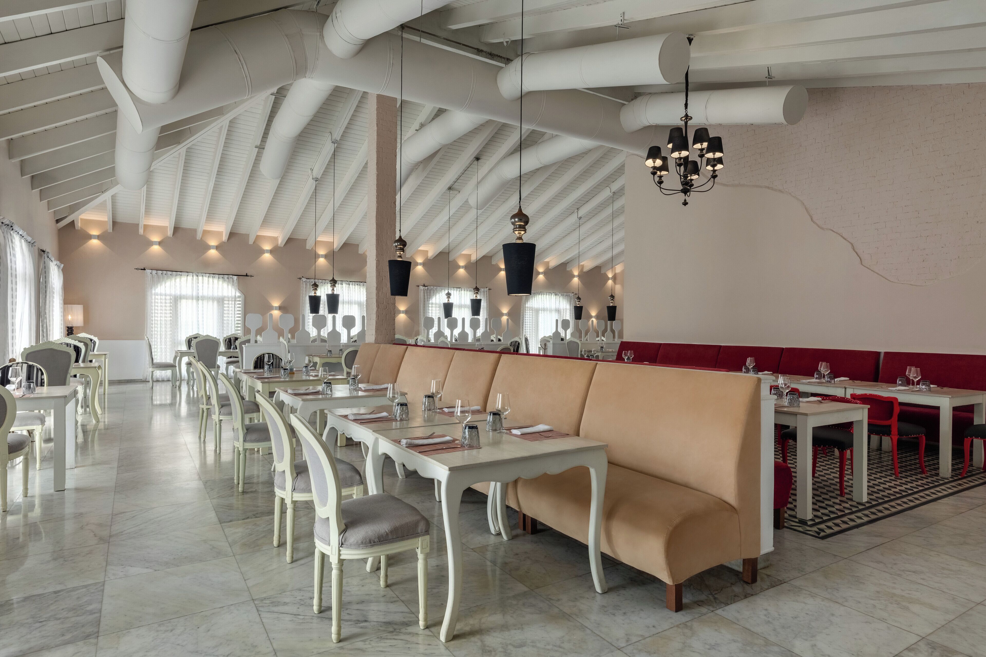 Uma sala de jantar ampla, com pé-direito alto, tubulações aparentes e uma combinação de assentos brancos e vermelhos. As mesas estão montadas com copos e talheres.