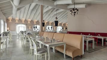 Uma sala de jantar ampla, com pé-direito alto, tubulações aparentes e uma combinação de assentos brancos e vermelhos. As mesas estão montadas com copos e talheres.