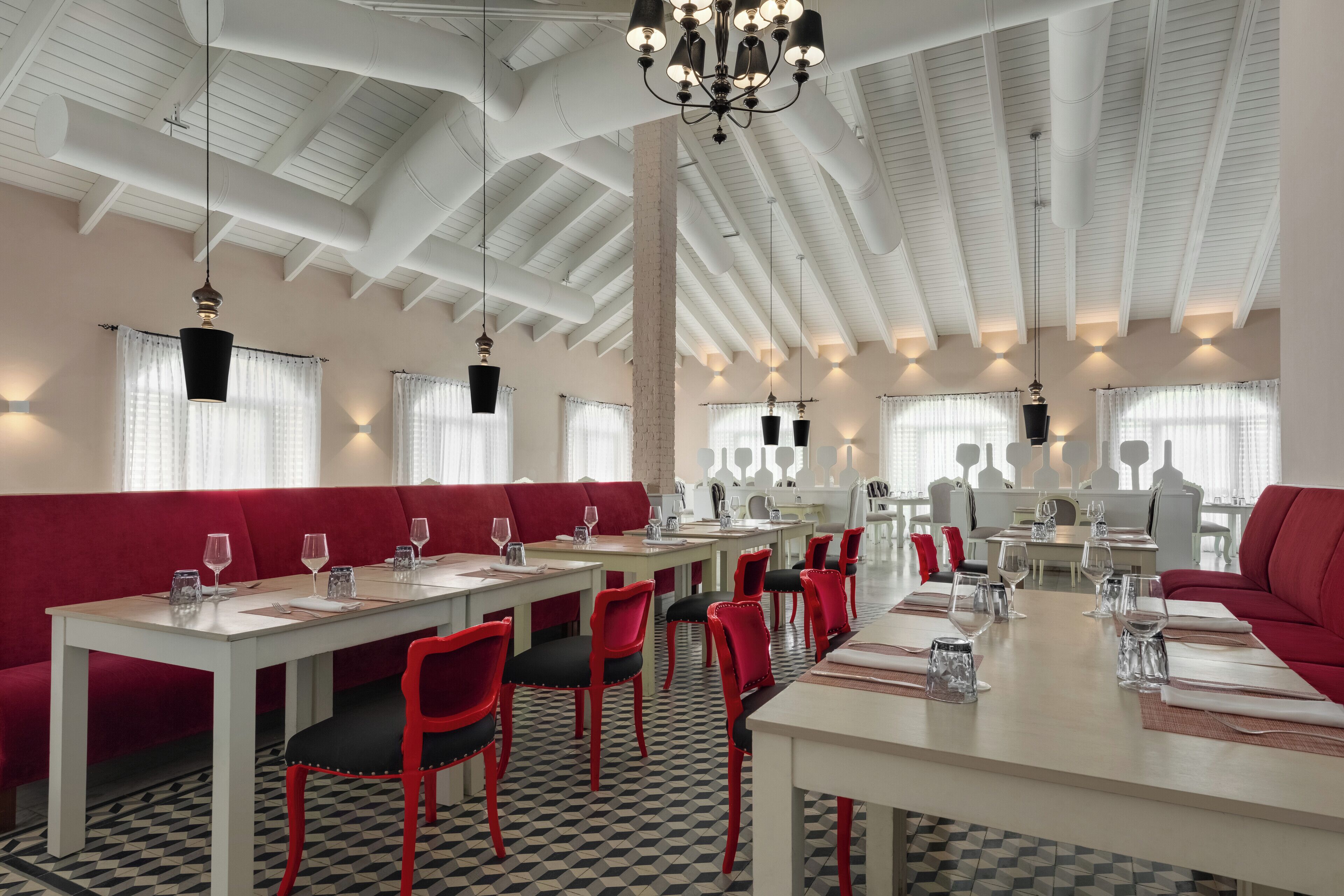 Restaurante com pé-direito alto, paredes brancas e chão quadriculado. Mesas dispostas com cadeiras vermelhas e toalhas de mesa brancas.