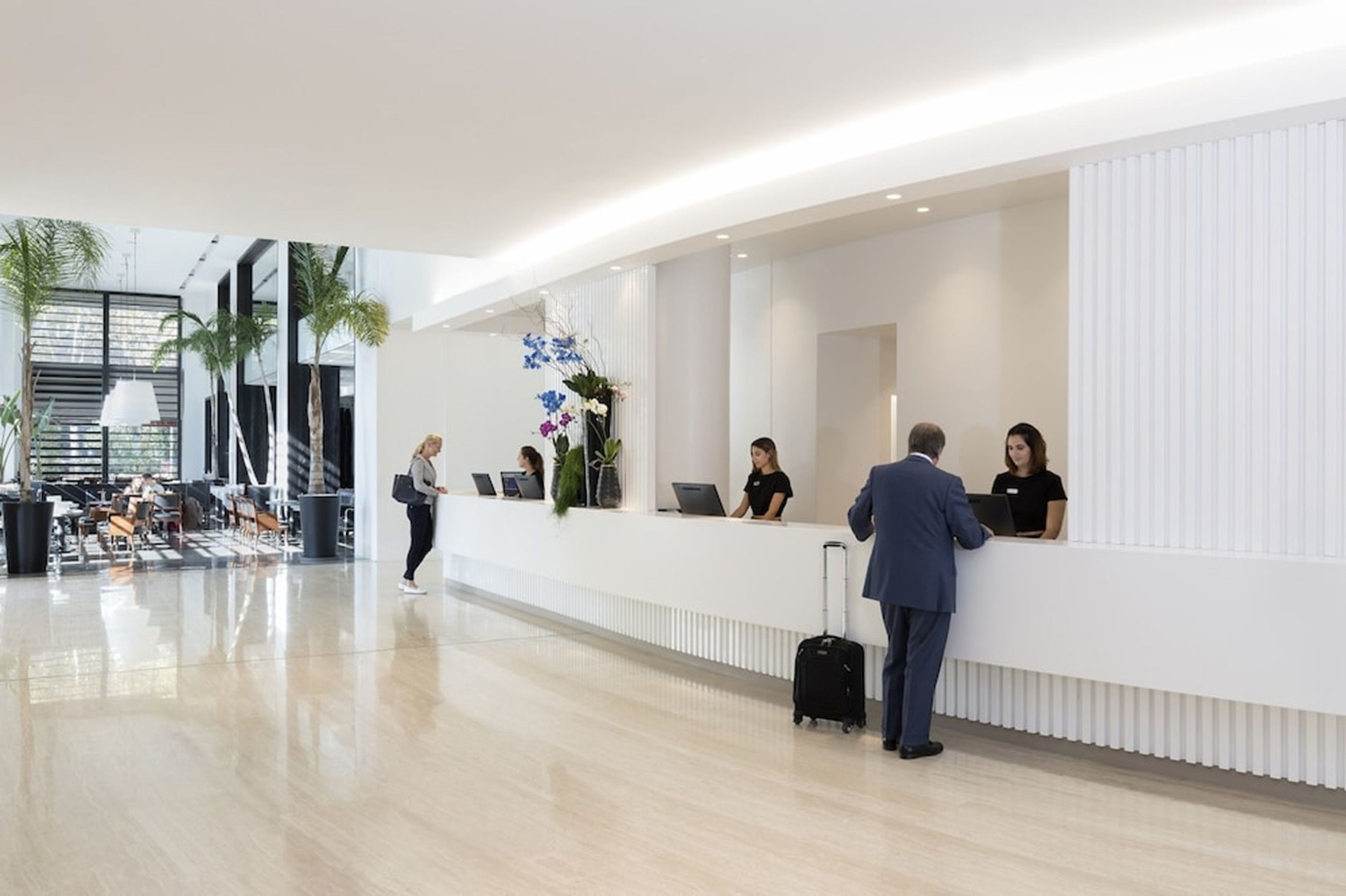 Uma área de recepção moderna com um balcão branco, vários funcionários e um homem com mala.