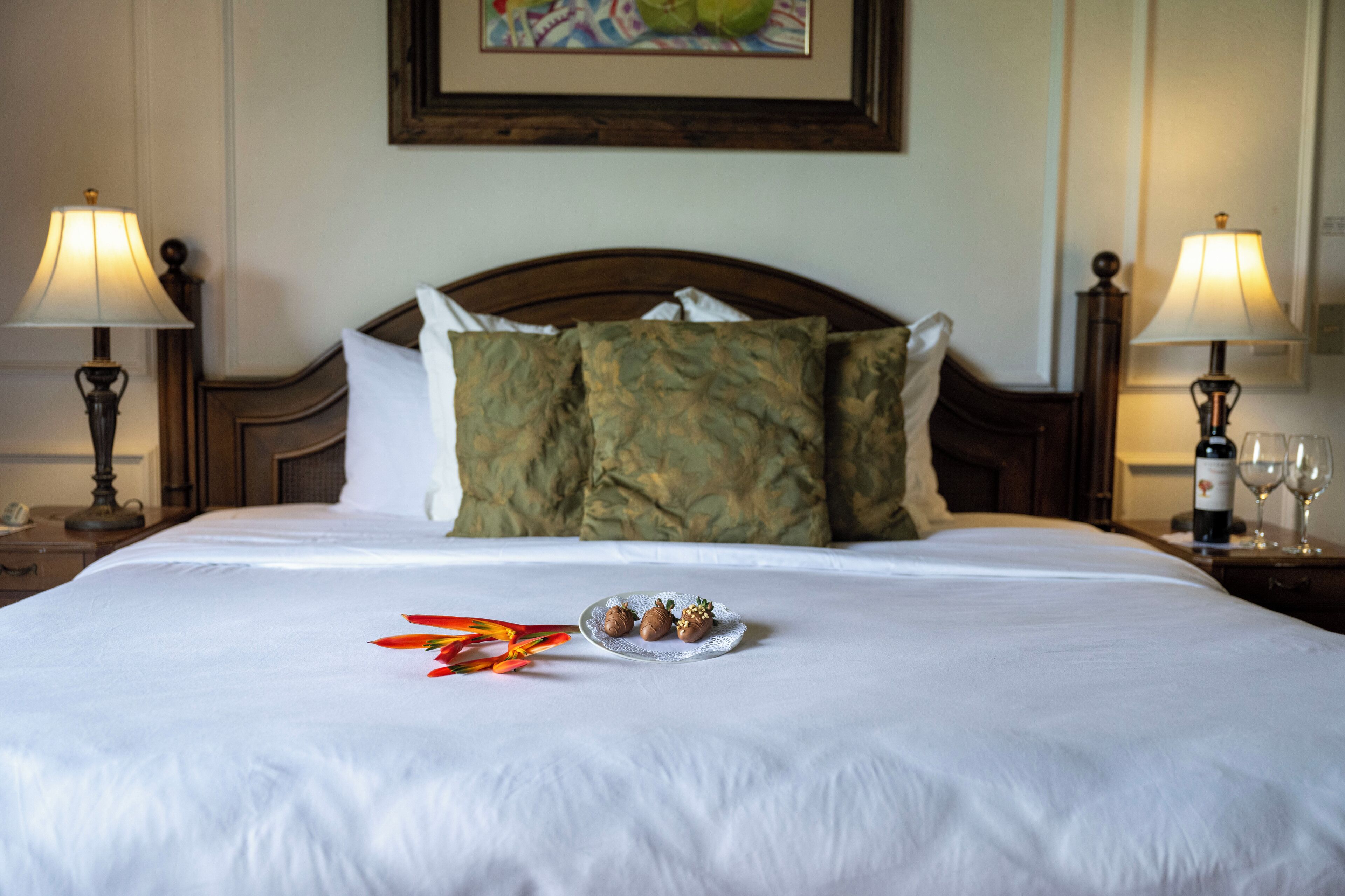 Uma cama bem arrumada com colcha branca, duas luminárias de cada lado e um prato de comida sobre a cama.