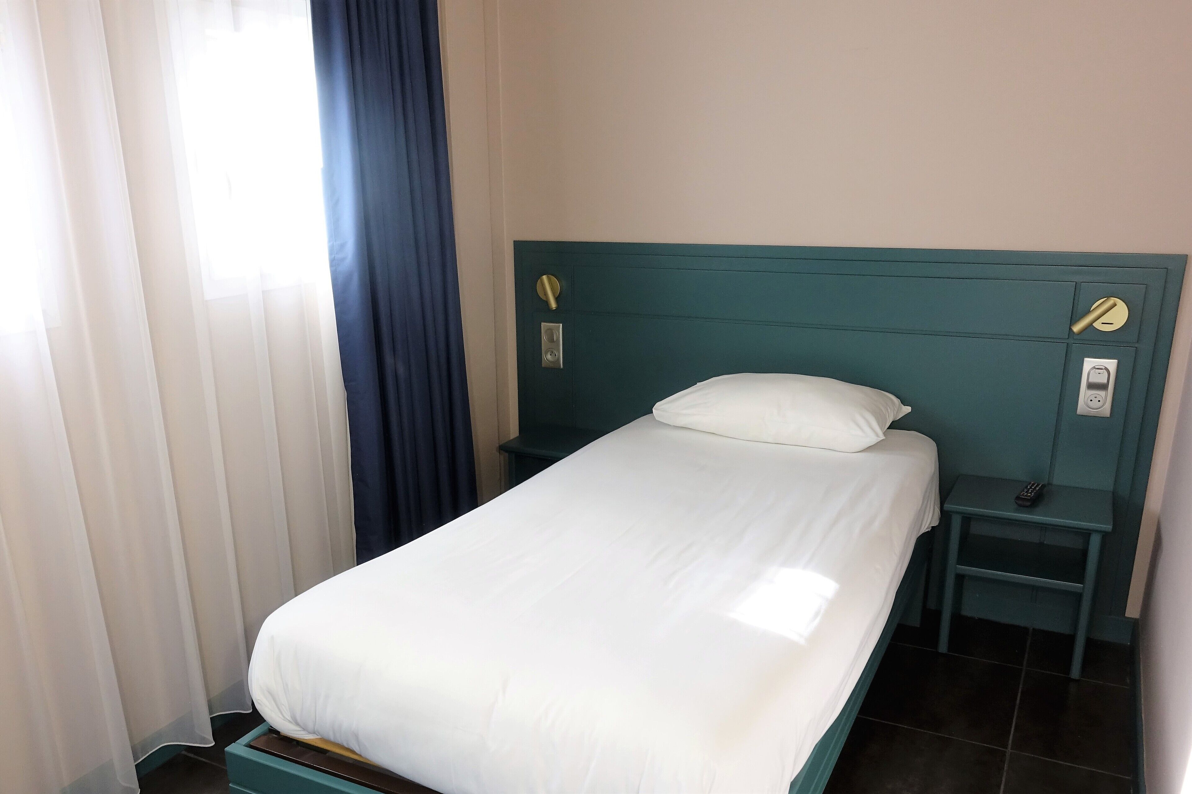 Uma cama de solteiro com colchão branco e cabeceira azul-esverdeada, uma mesa de cabeceira com controle remoto e uma janela com cortinas leves.