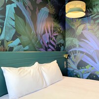 Cama com roupa de cama branca e cabeceira verde em quarto com papel de parede de padrão tropical.