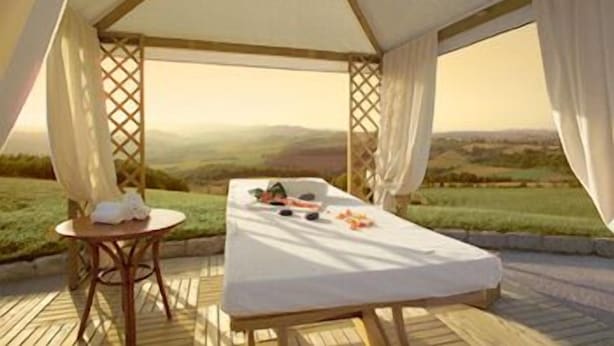Uma área relaxante ao ar livre com tendas, mesa de massagem, toalhas e uma mesa com garrafa e frutas.
