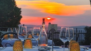 Mesa posta para refeição com taças de vinho, garrafa e castiçais, com o pôr do sol ao fundo.