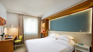 Quarto de hotel com uma cama grande, uma escrivaninha com cadeira, mesa de cabeceira e luminária na parede.