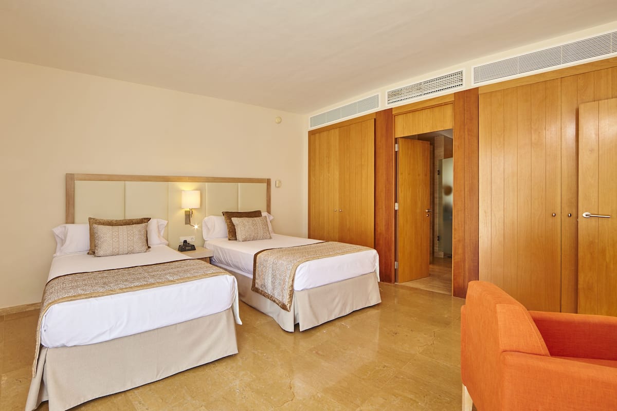 Quarto de hotel com duas camas, mesa de cabeceira, luminária, armário de madeira e uma poltrona vermelha.