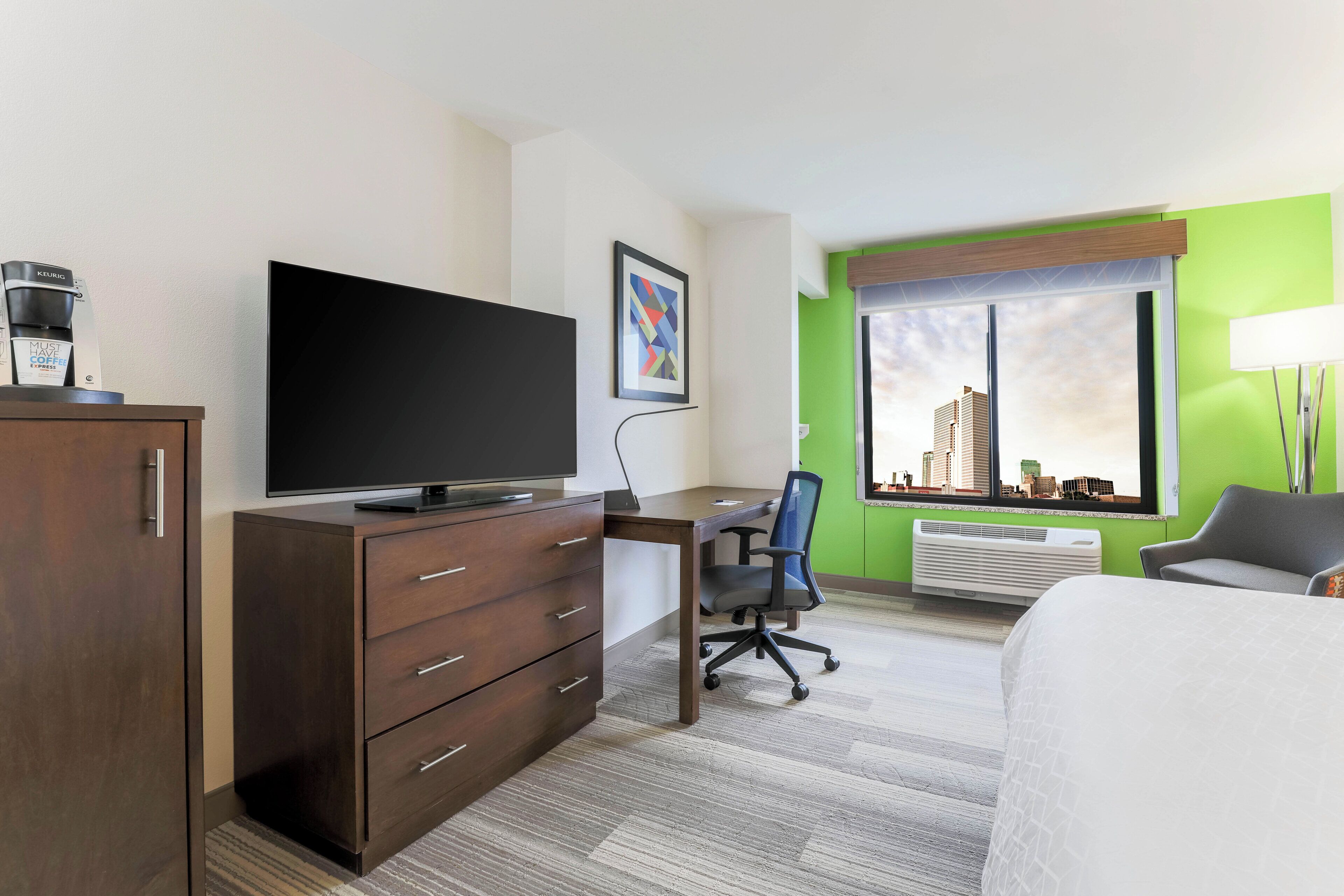 Quarto de hotel com televisão de tela plana, escrivaninha, cadeira, cama, guarda-roupa, quadro decorativo, janela com vista para a cidade e luminária.